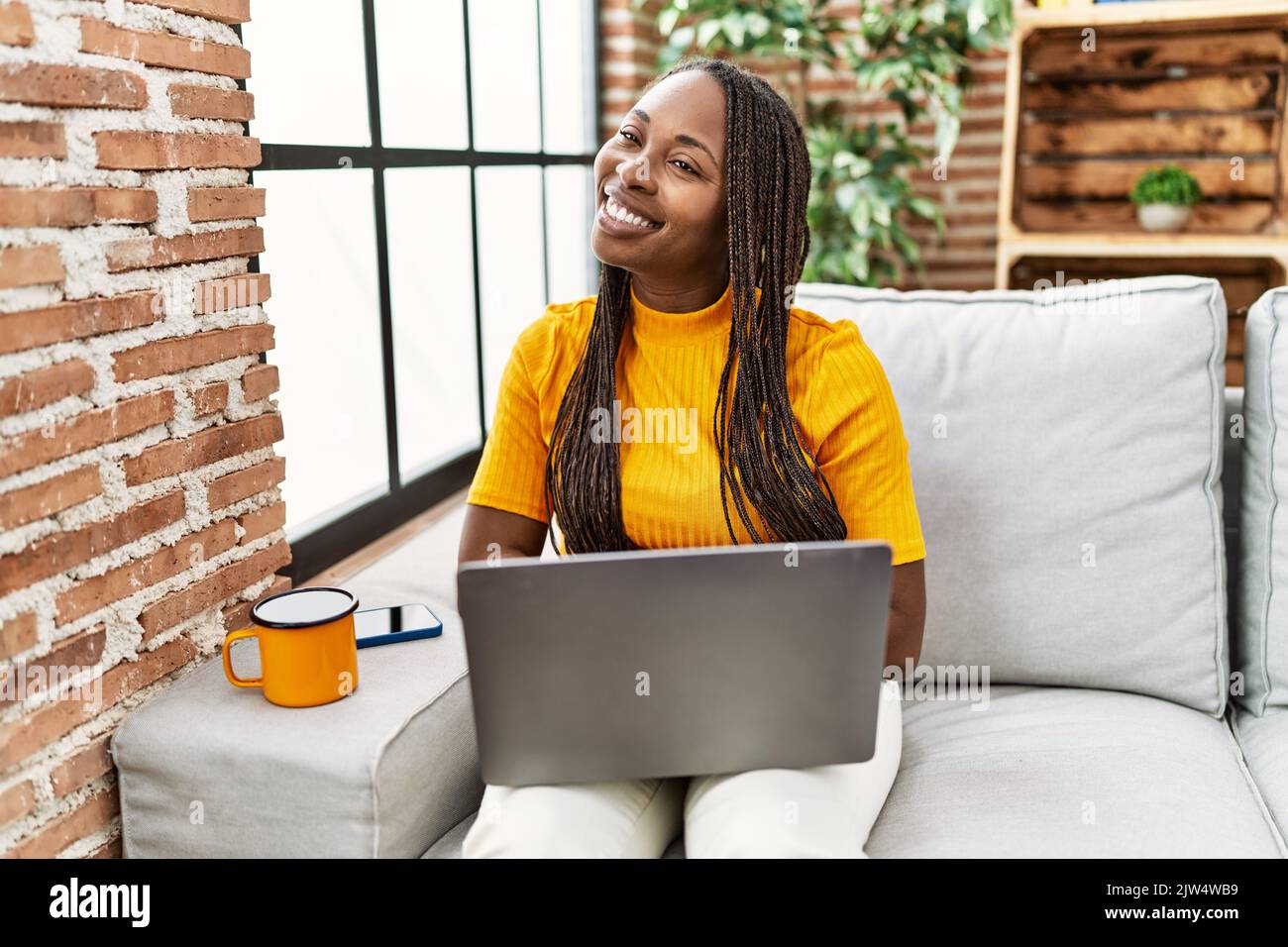 African woman sitting on the sofa using laptop at home looking away to ...