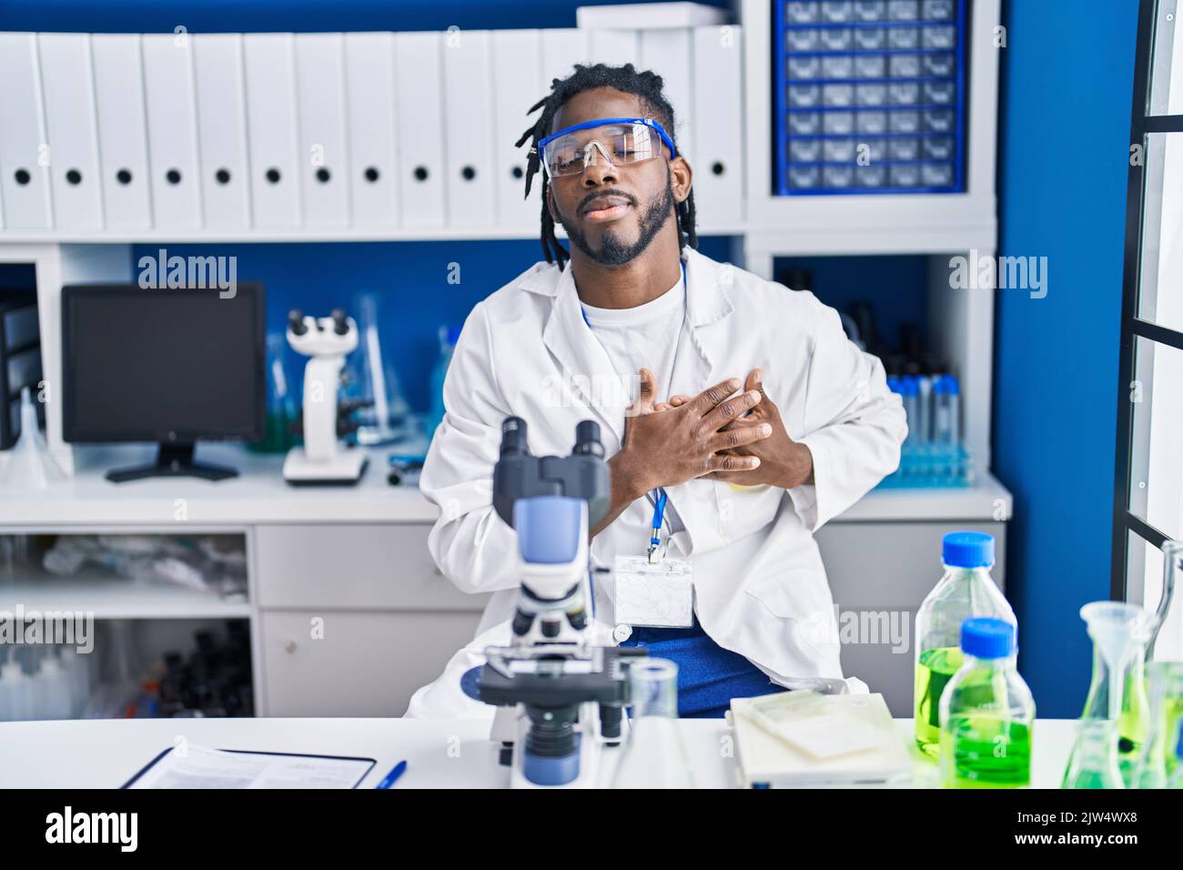 African man with dreadlocks working at scientist laboratory smiling with hands on chest, eyes ...