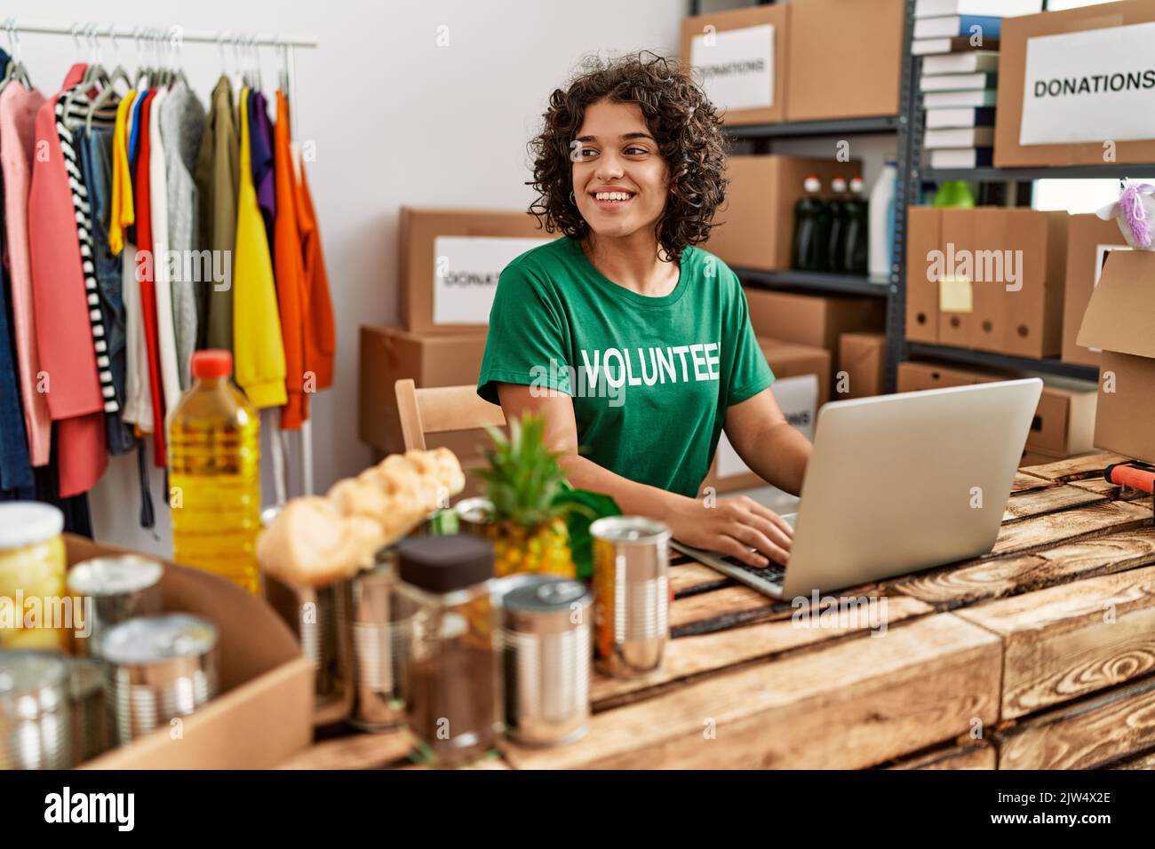 Young hispanic woman wearing volunteer uniform using laptop at charity ...