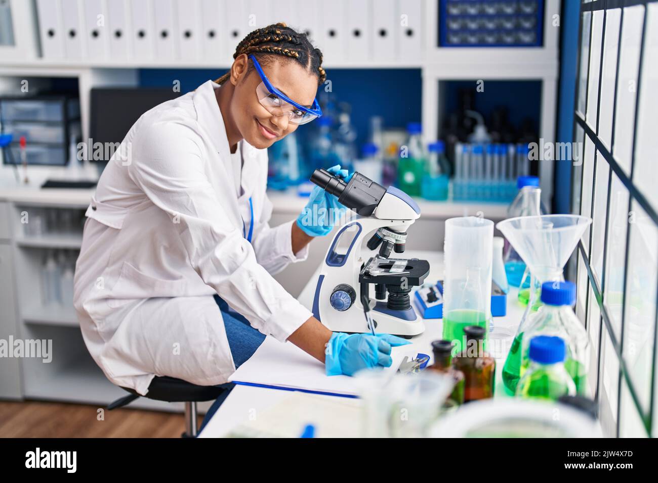 African american woman scientist using microscope writing document at ...