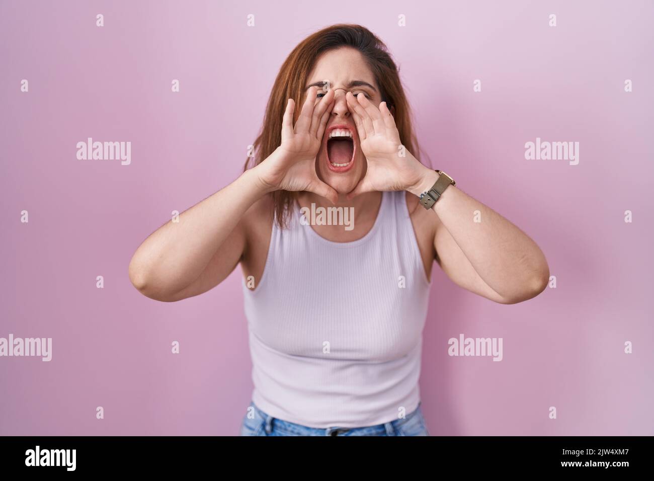 Brunette woman standing over pink background shouting angry out loud ...