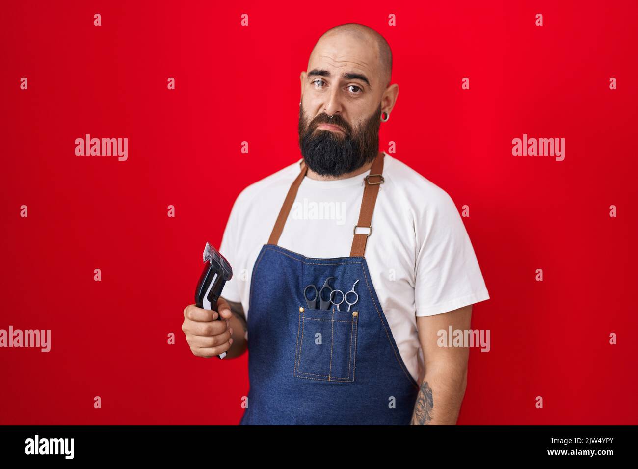 Young hispanic man with beard and tattoos wearing barber apron holding ...