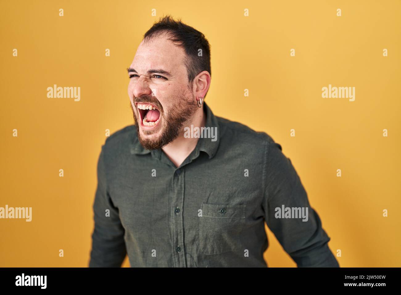 Plus size hispanic man with beard standing over yellow background angry ...