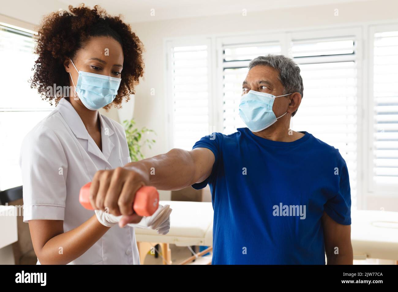 Mixed race female physiotherapist wearing mask helping senior exercise ...