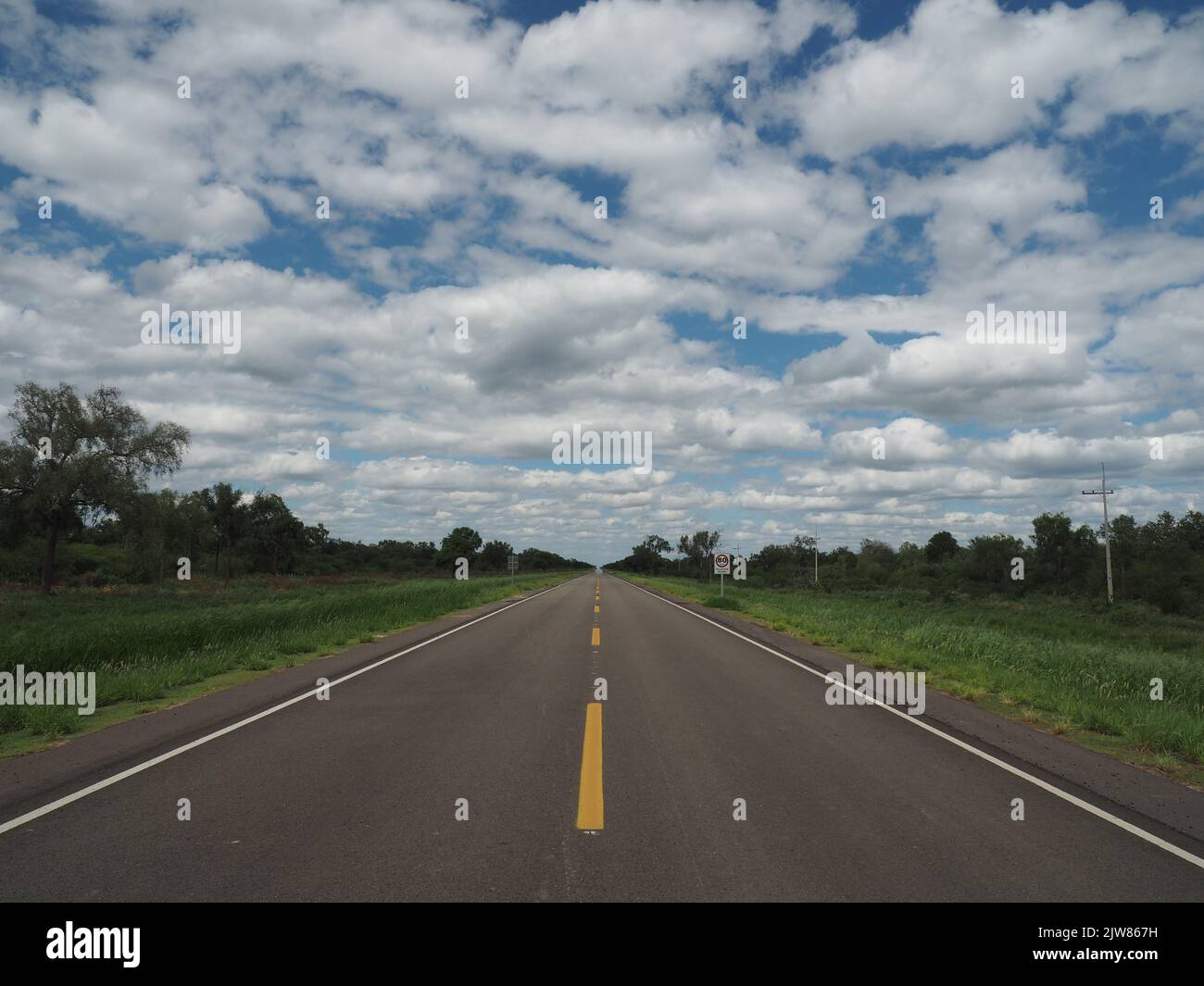 A wide road surrounded by rees and bushes under the blue cloudy sky ...
