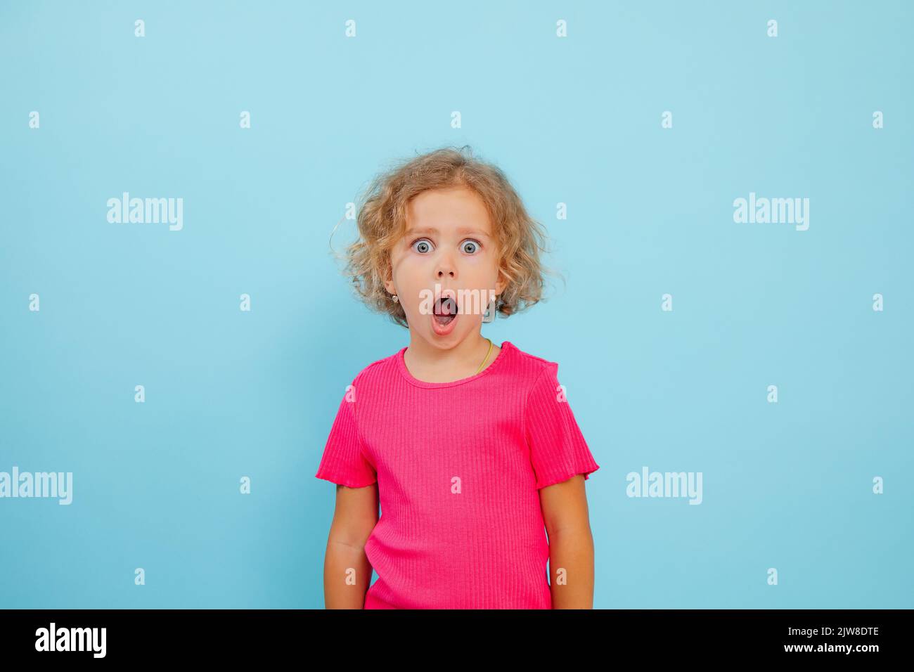 Little expressive, shocked, emotional curly blonde girl in pink shirt with open mouth, look at ...