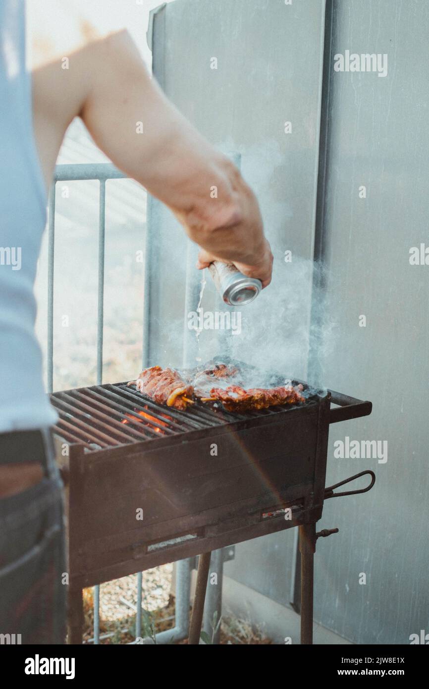A vertical shot of a hand pouring a can of water on meat on a grill ...