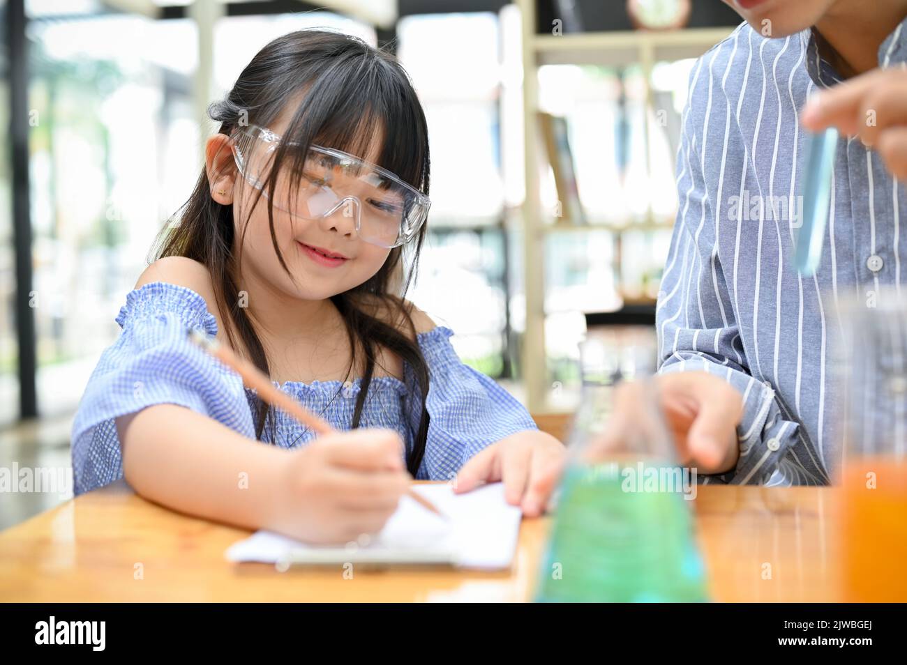 Cute young Asian girl in science goggles taking notes in her notebook ...