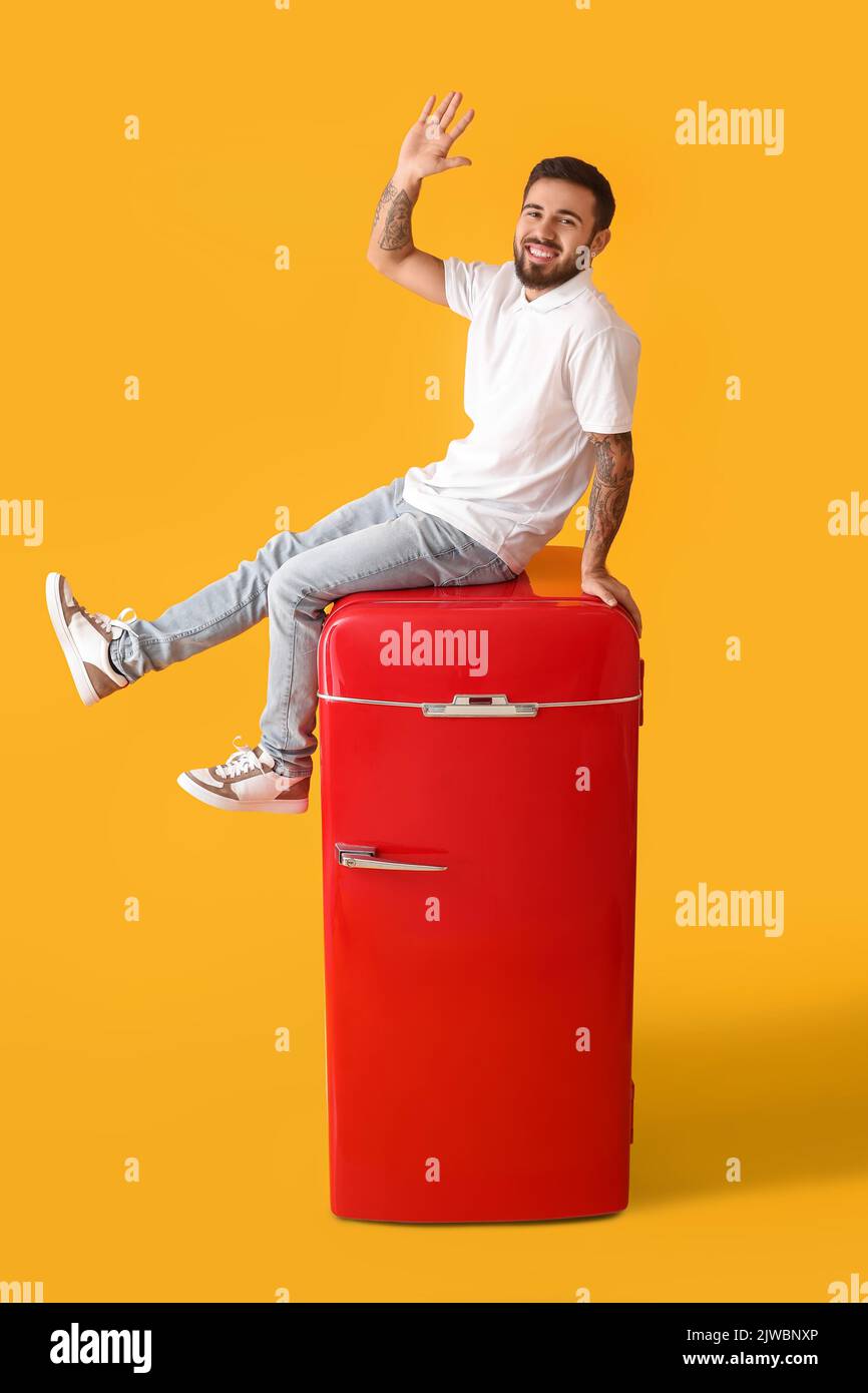 Cool tattooed young man sitting on red fridge against yellow background ...