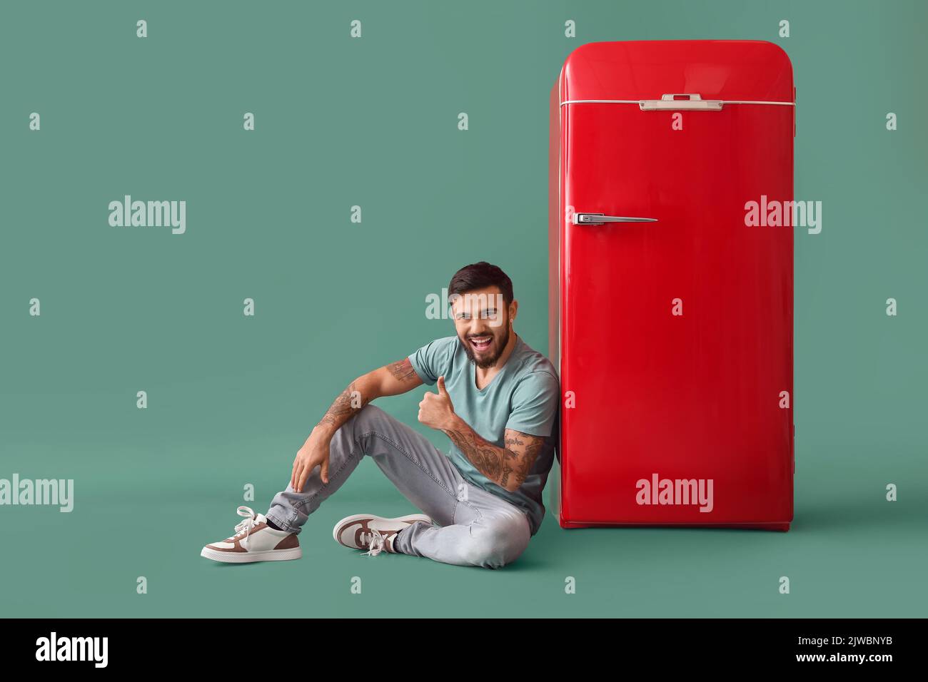 Cool tattooed young man sitting near red fridge on green background ...