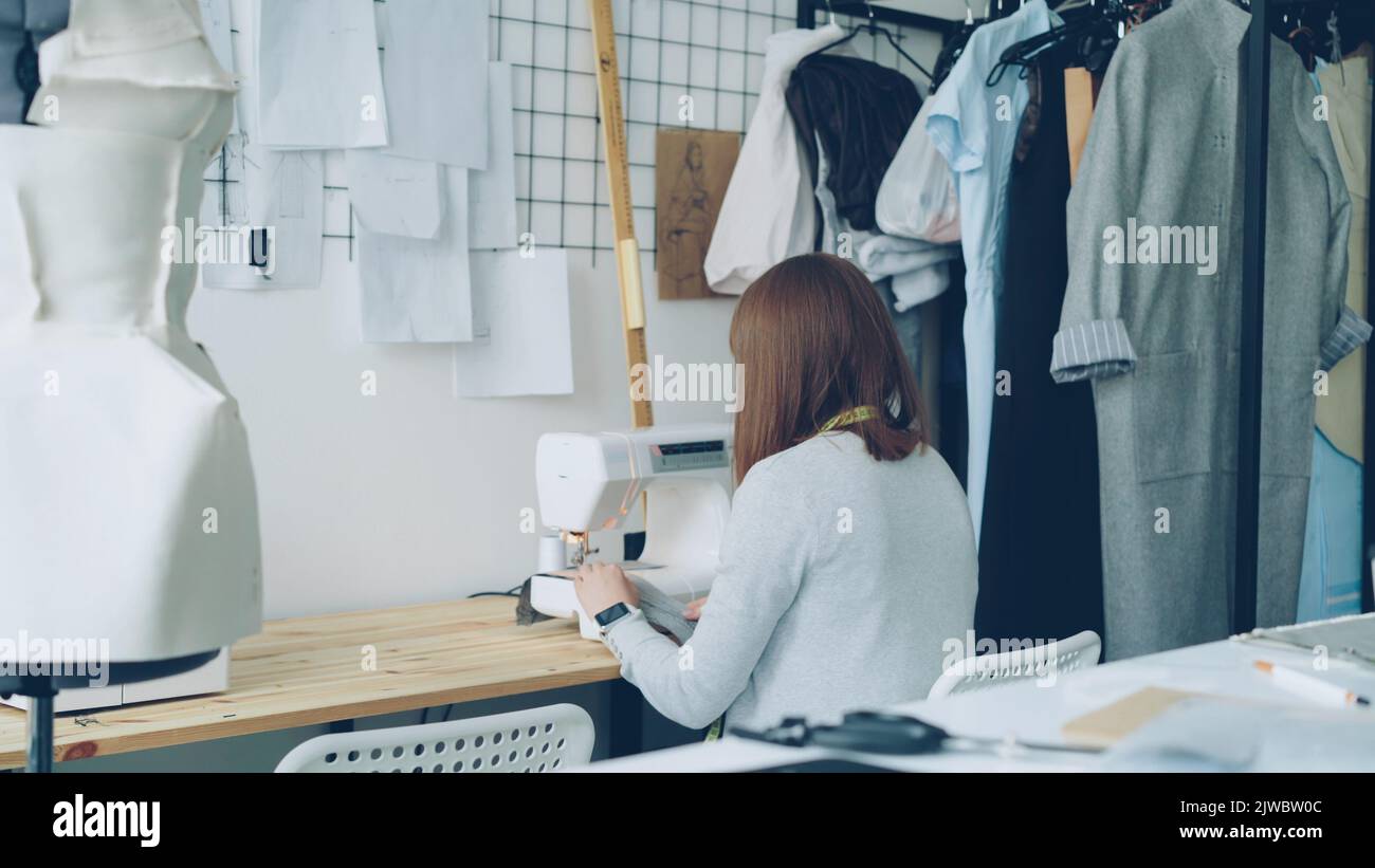 Back view of young woman clothing designer working with sewing machine ...