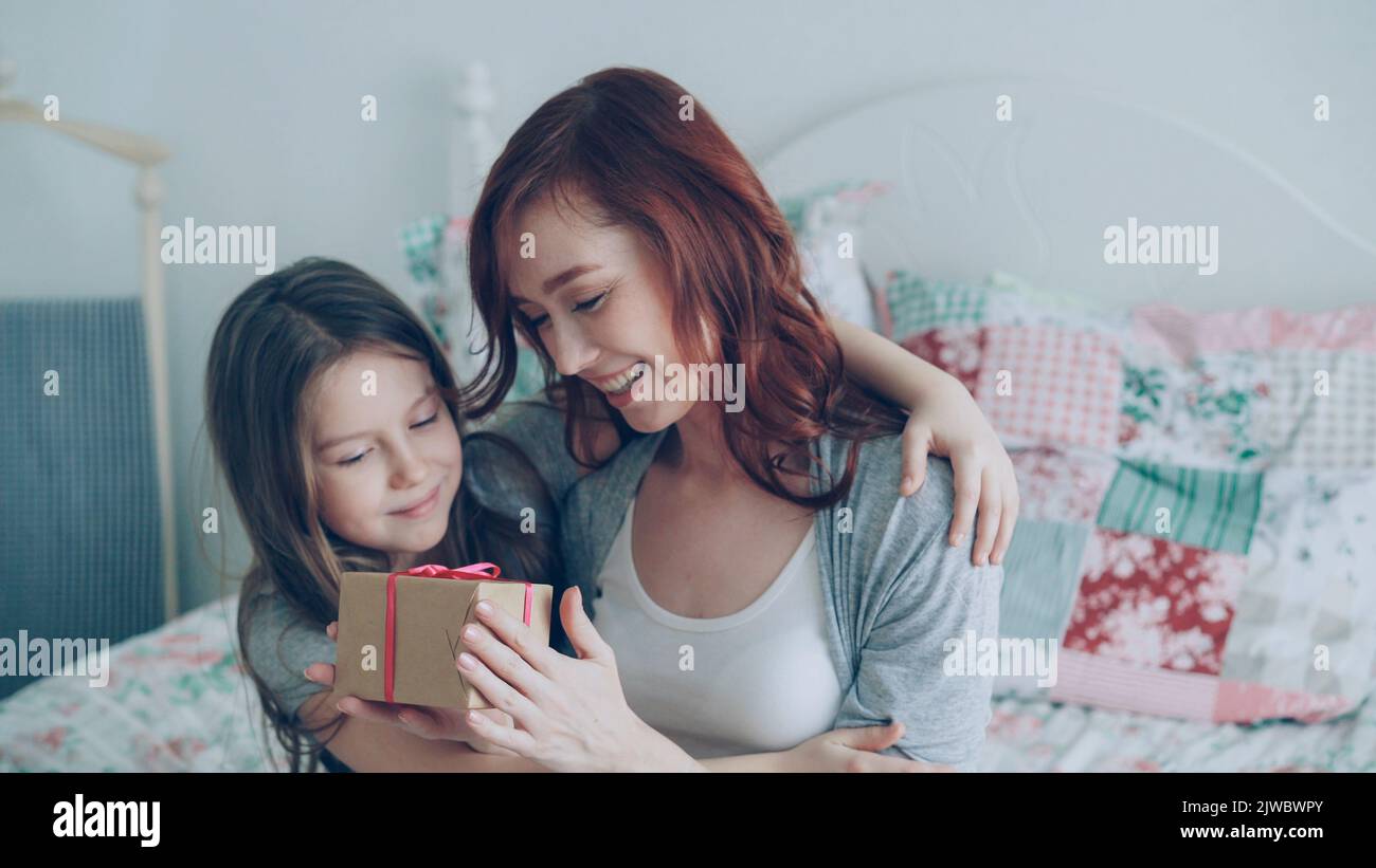 Little cute girl giving gift box to her young happy mom celebrating mothers day sitting on bed ...