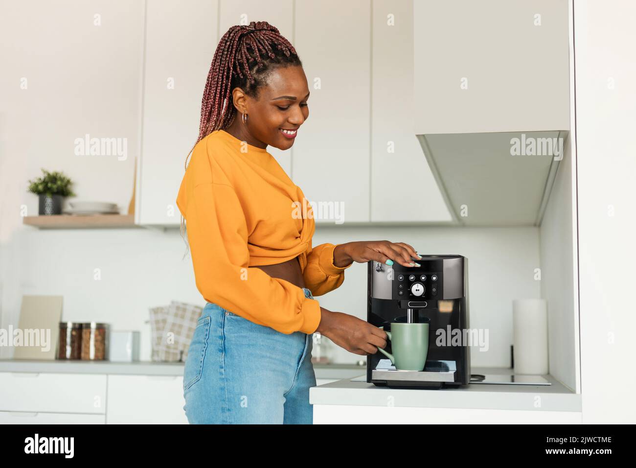 Excited young black woman making fresh aromatic coffee in modern machine in kitchen interior ...