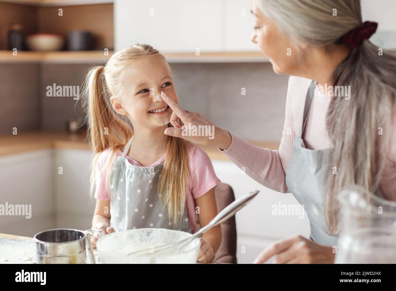 Happy senior woman in apron touches nose of little girl, make dough and ...