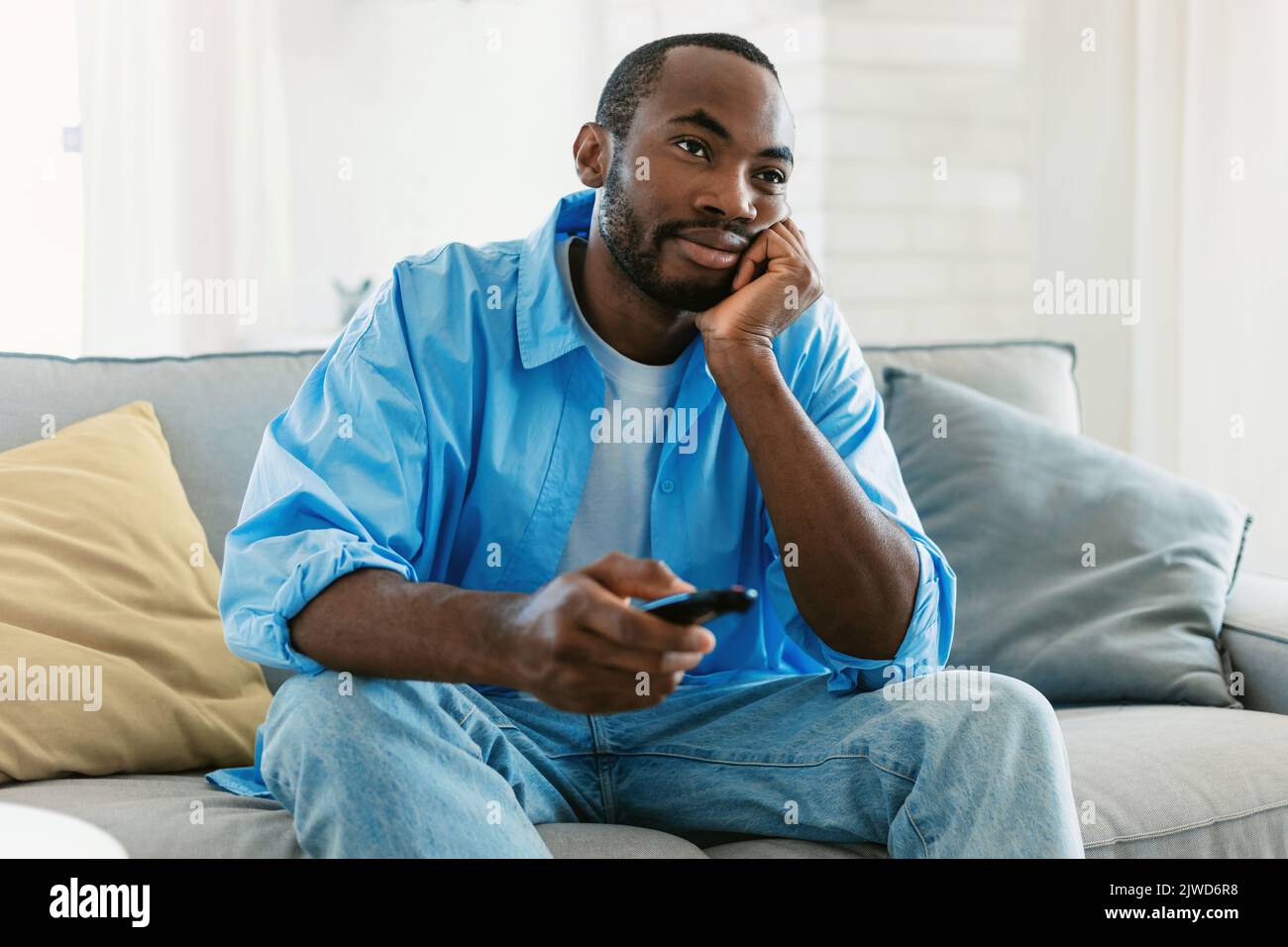 Bored black man watching boring TV, switching channels holding remote controller, sitting on ...