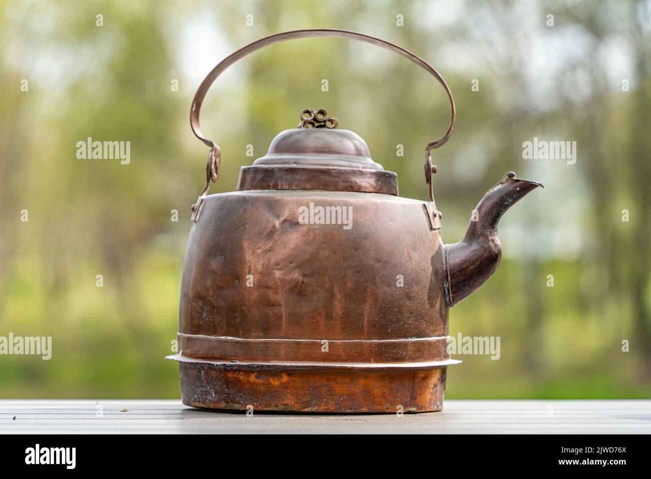 Old copper kettle on the wooden table with trees at the background ...