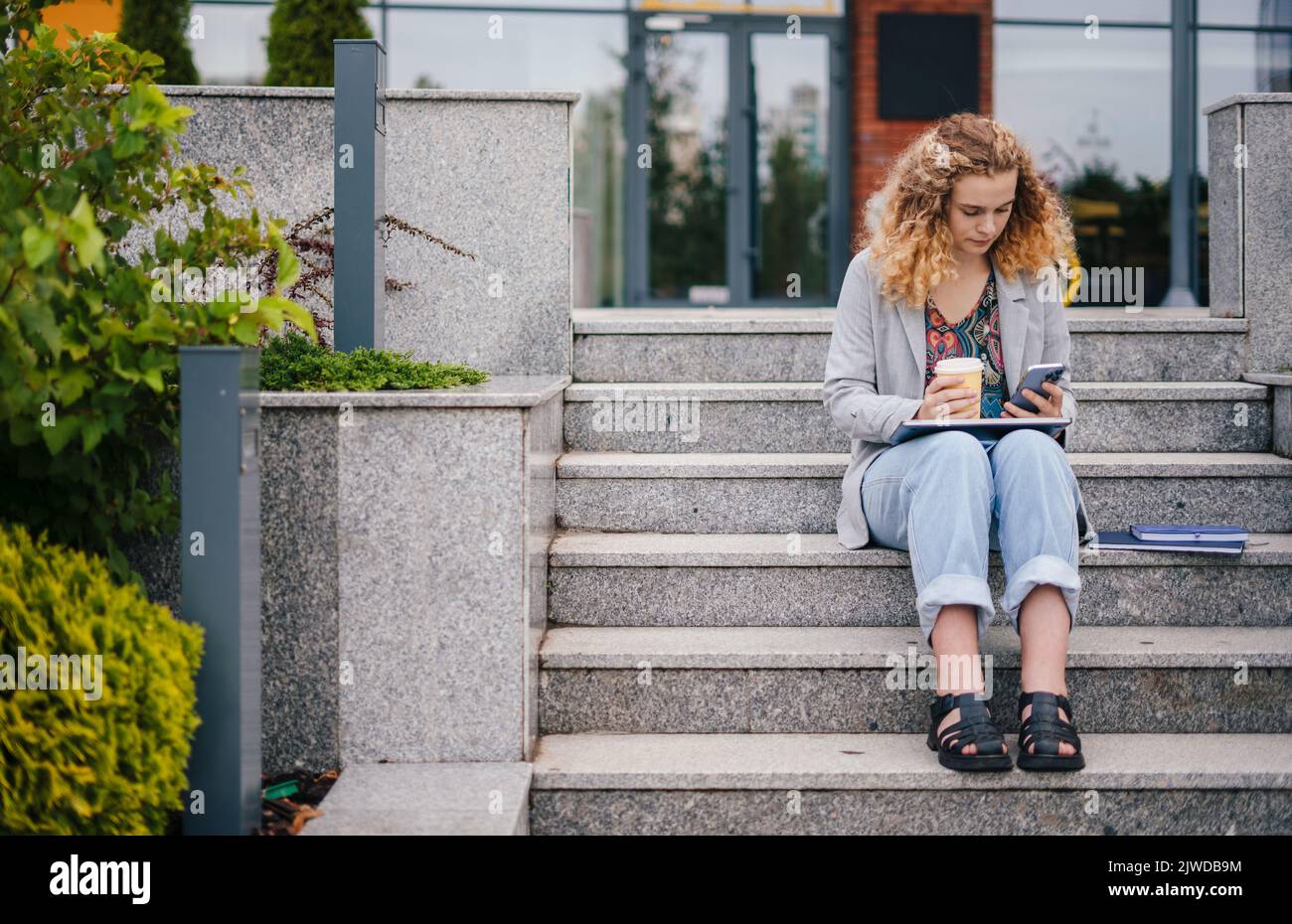 Caucasian student with coffee to go using mobile phone, sitting on ...