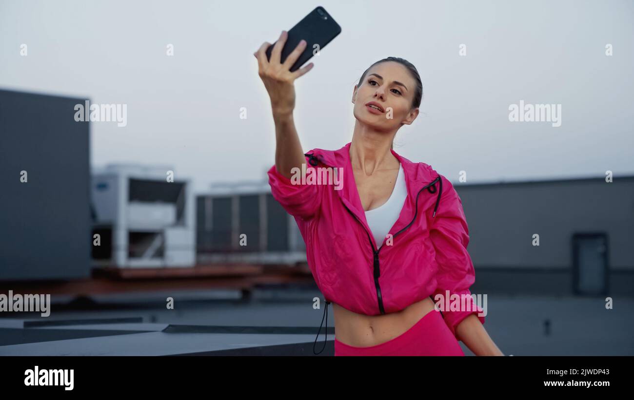 Sportswoman in pink sportswear taking selfie on smartphone on roof of ...