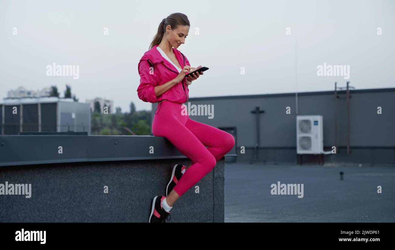 Smiling sportswoman using smartphone on roof of building in evening ...