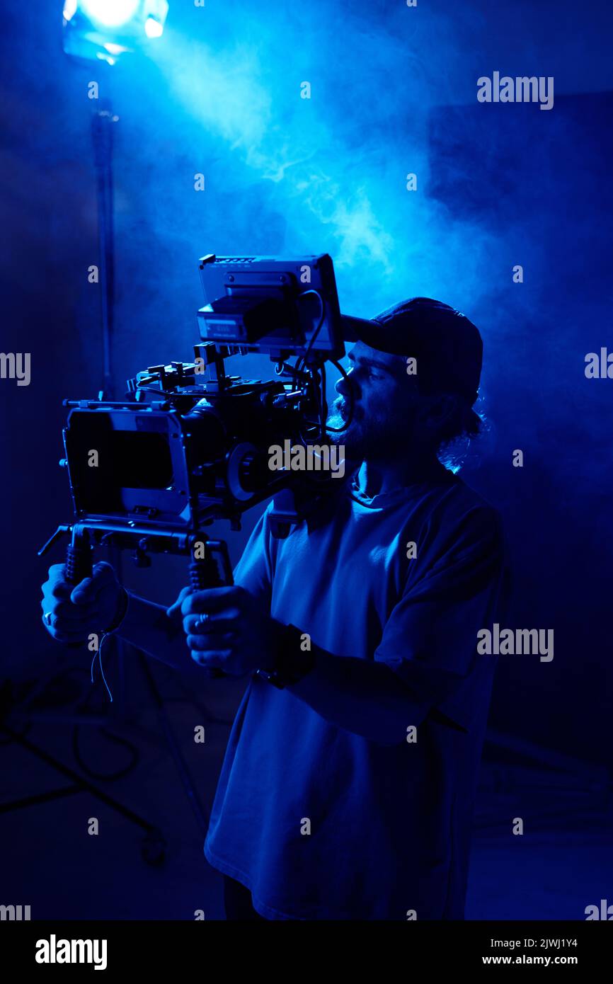 Serious young cameraman with steadicam standing against spotlight in ...