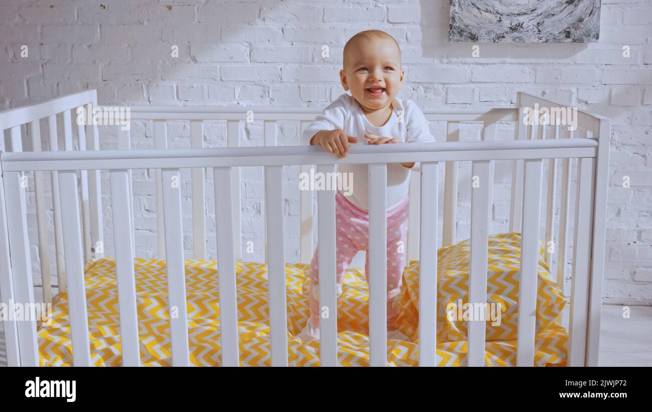 happy infant child standing in baby crib and smiling near brick wall ...