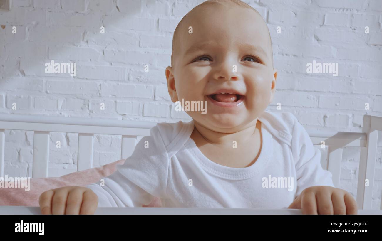 cheerful baby standing in white baby crib while smiling at home Stock ...