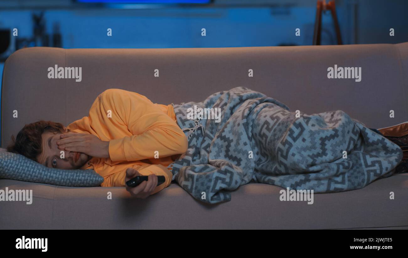 sleepy man in yellow hoodie lying under blanket and watching tv on sofa ...
