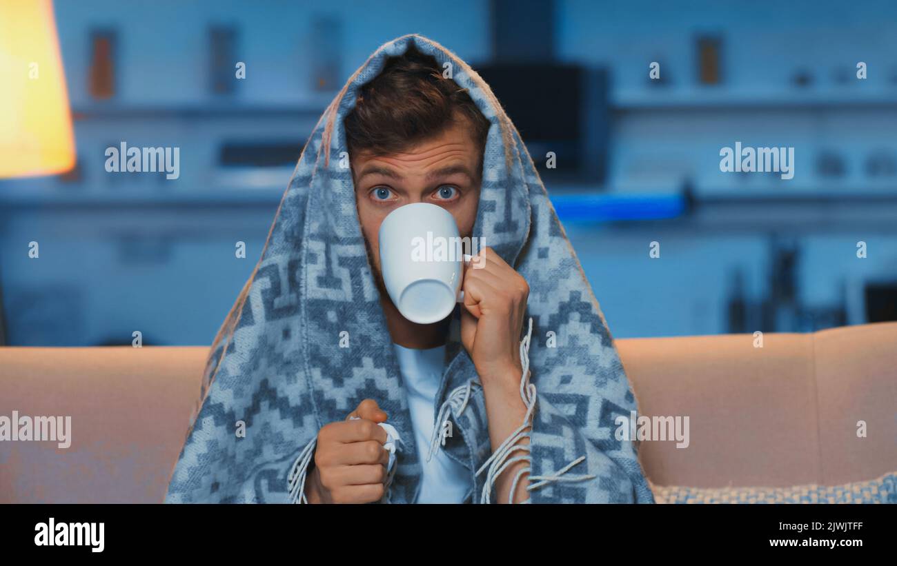 sick man with blue eyes drinking tea while sitting under blanket in ...