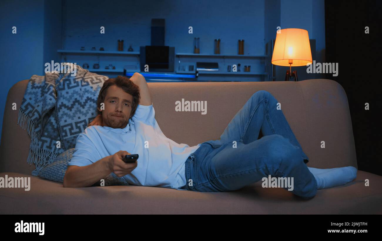 bored young man lying on couch and watching tv in modern living room at ...