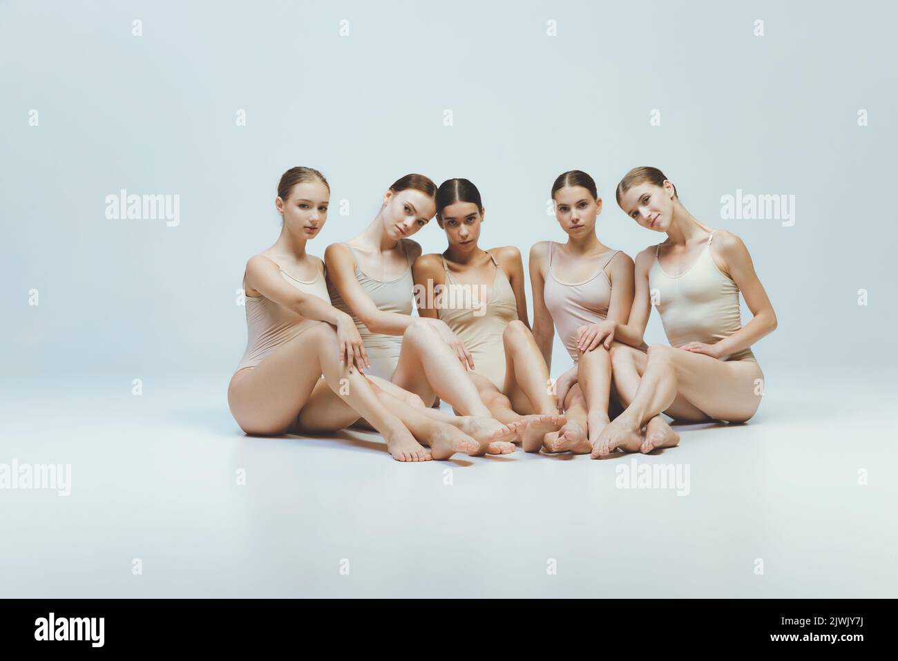 Group of young girls, ballet dancers in beige bodysuits sitting together, posing isolated over ...