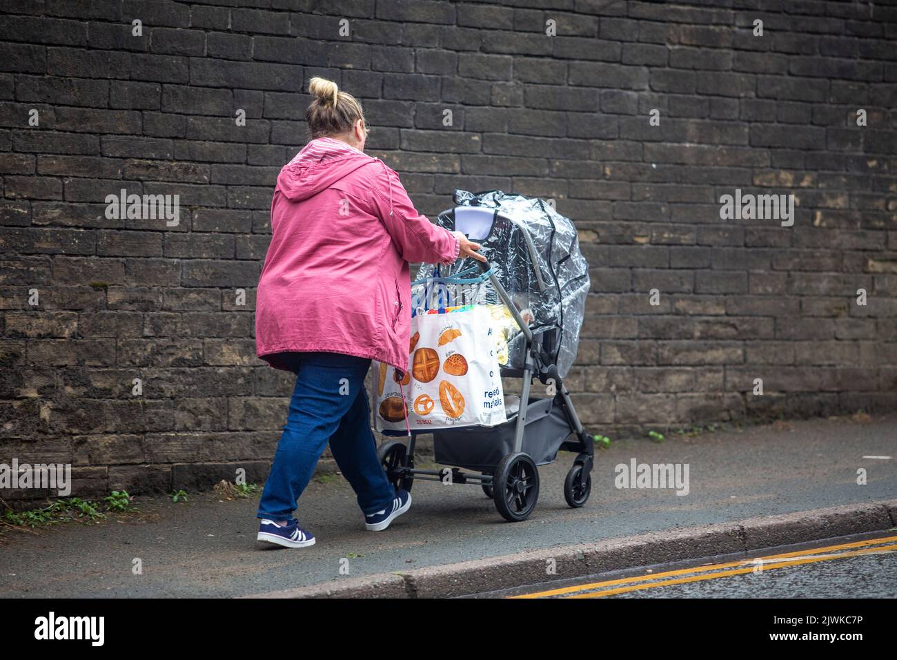 A lady with a Tesco shopping bag pushes a baby in a pram on grey wet ...