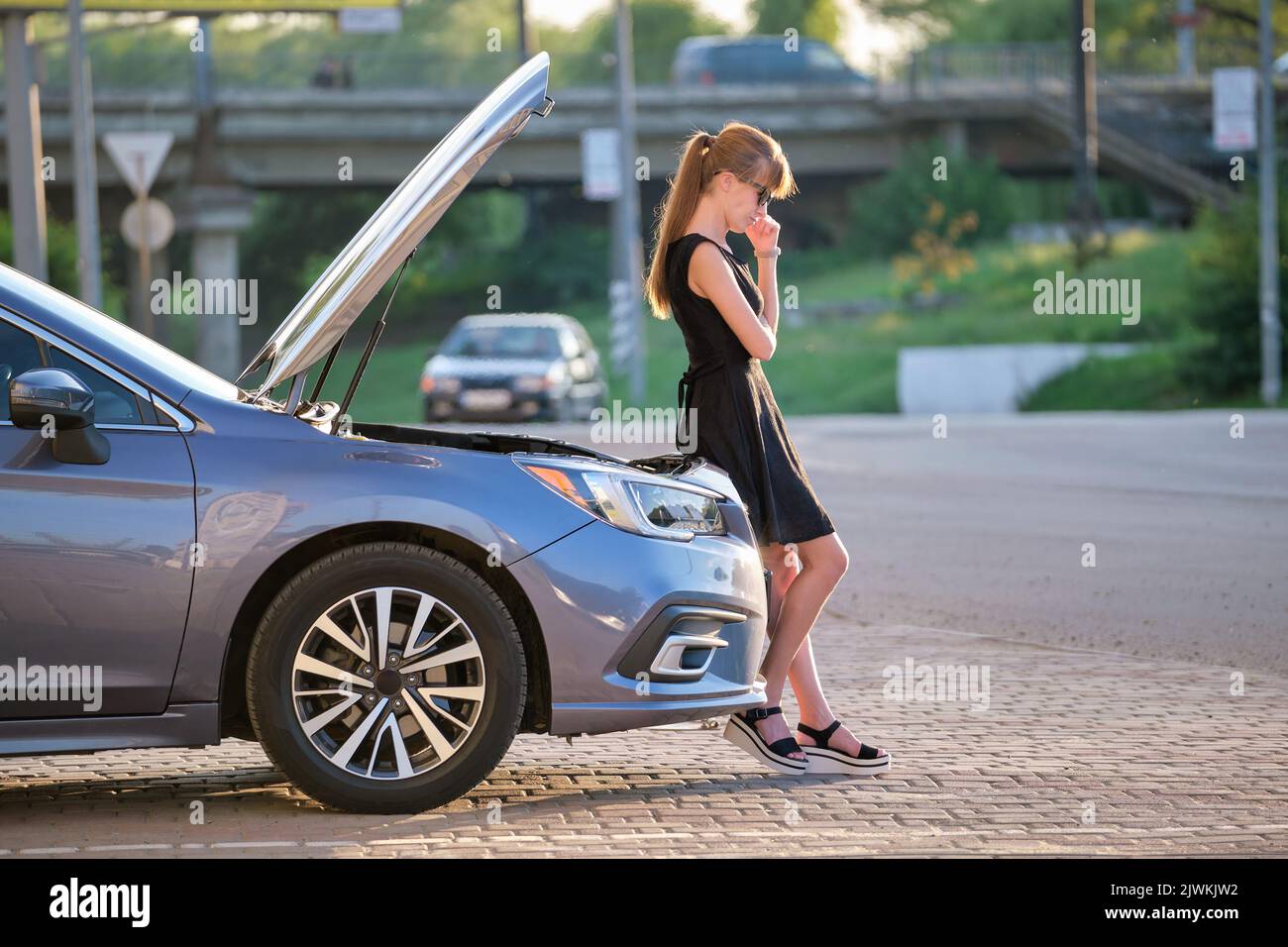 Sad female driver standing on a city street near her car with popped up ...