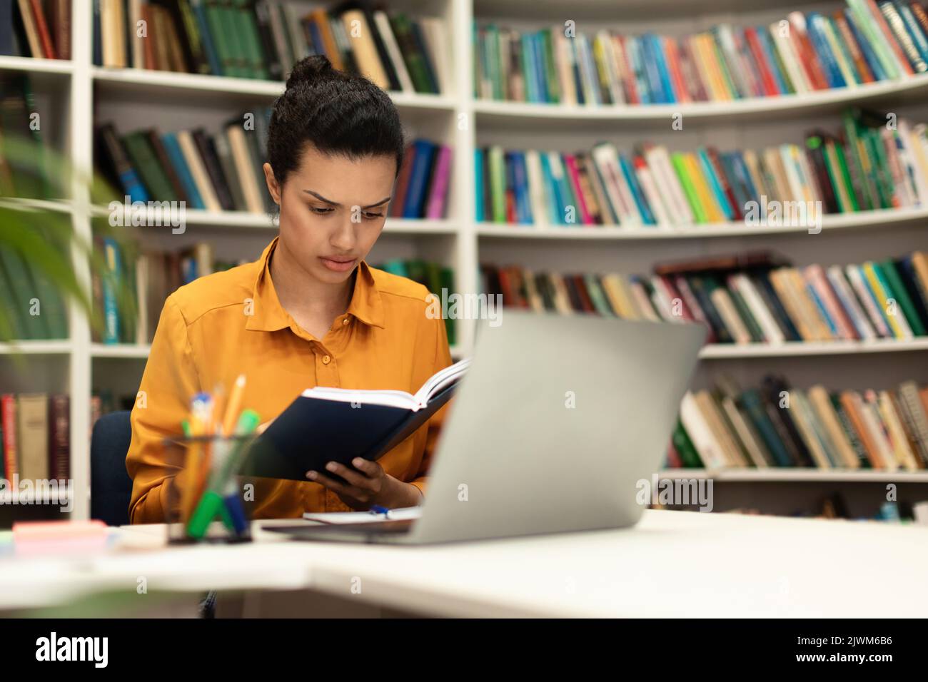 Concentrated mixed race woman reading book, sitting in library with laptop pc and study ...