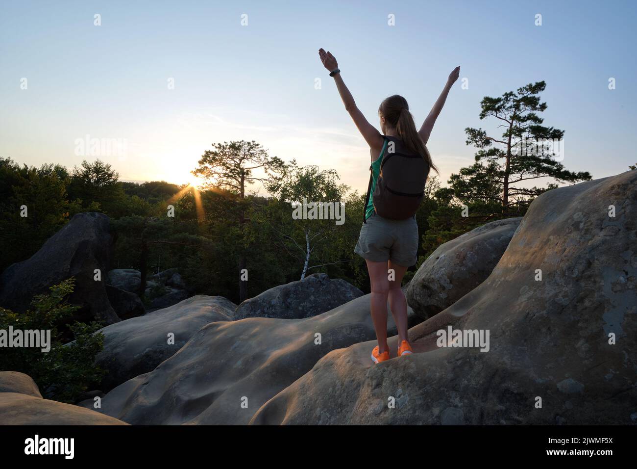 Sportive woman standing alone on hillside trail with raised up arms at sunset. Female hiker ...
