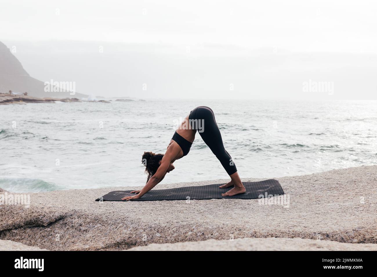 Side view of slim woman practicing Downward Facing Dog pose by ocean at ...
