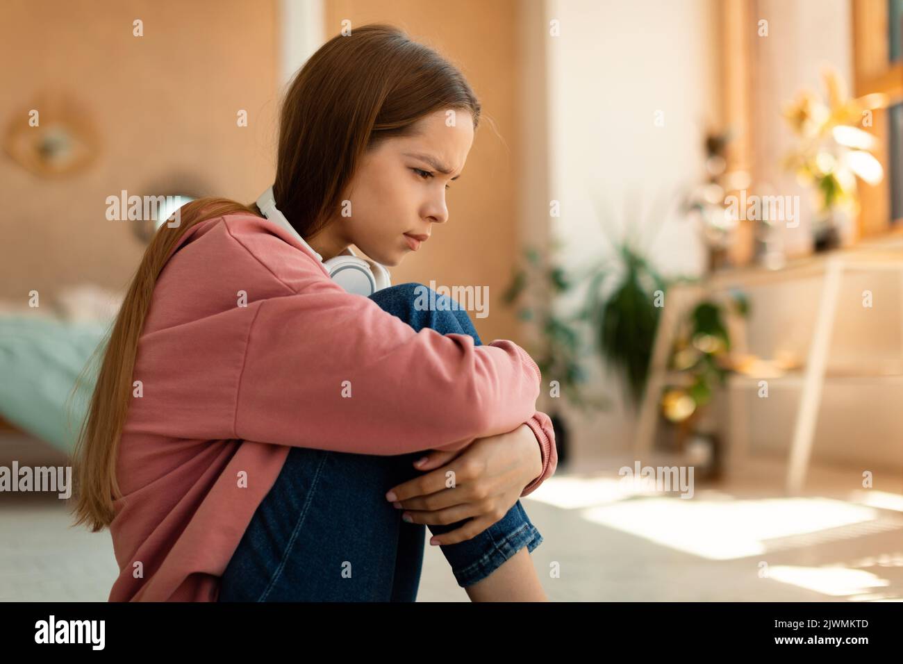 Teen depression concept. Stressed teenage girl sitting on floor at her bedroom, looking down ...
