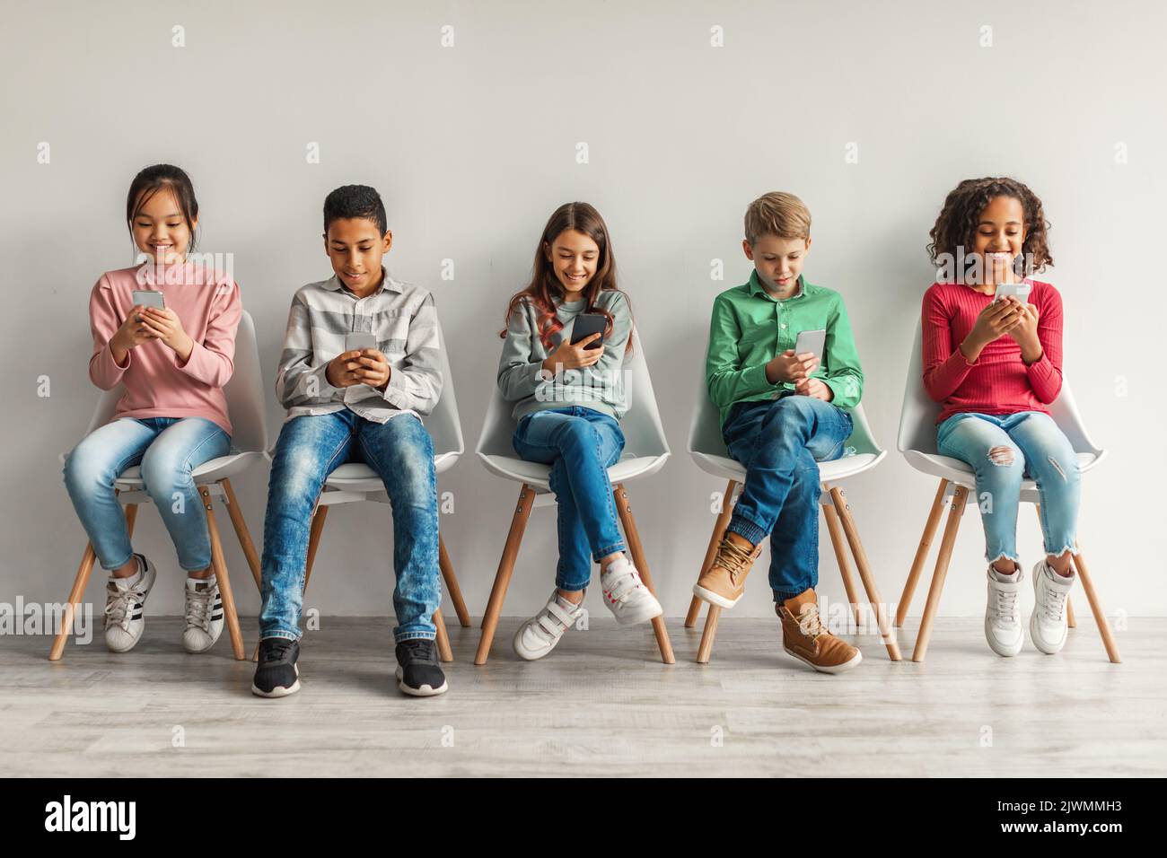 Multiracial Preteen Kids Using Cellphones Sitting Near Gray Wall Indoor ...