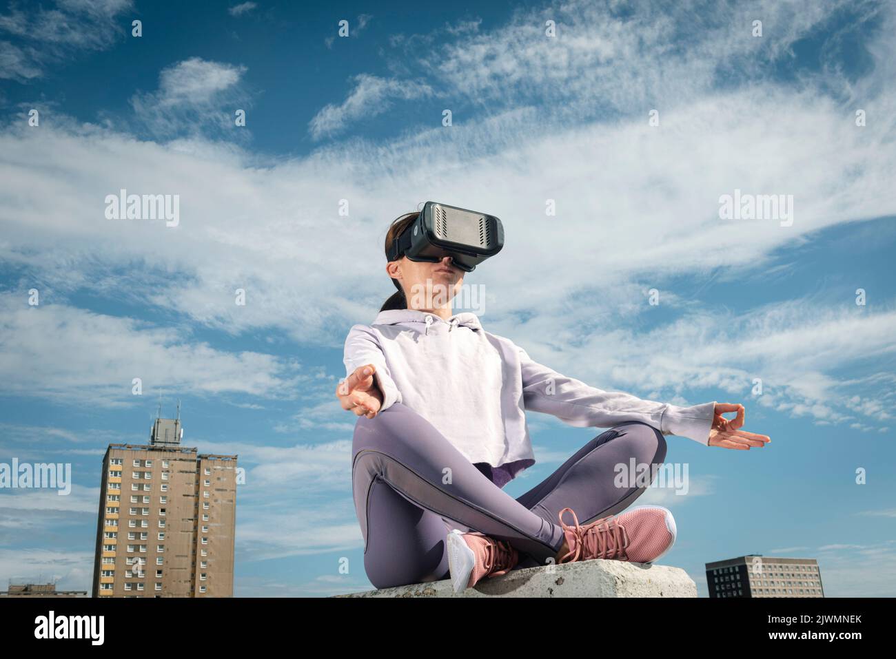 woman sitting wearing VR goggles and yoga meditating, outside, urban ...