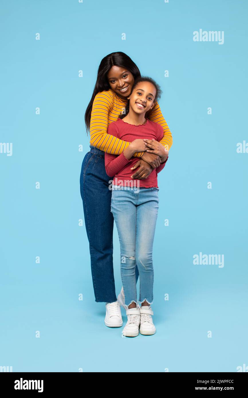 Happy black mother hugging her lovely daughter standing in studio over blue background, full ...