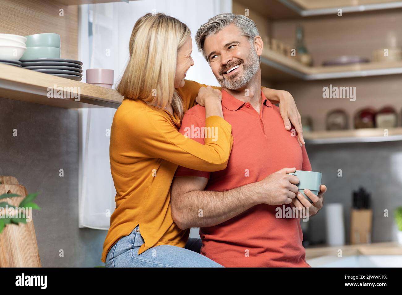 Loving lady hugging her smiling husband, drinking coffee at kitchen ...