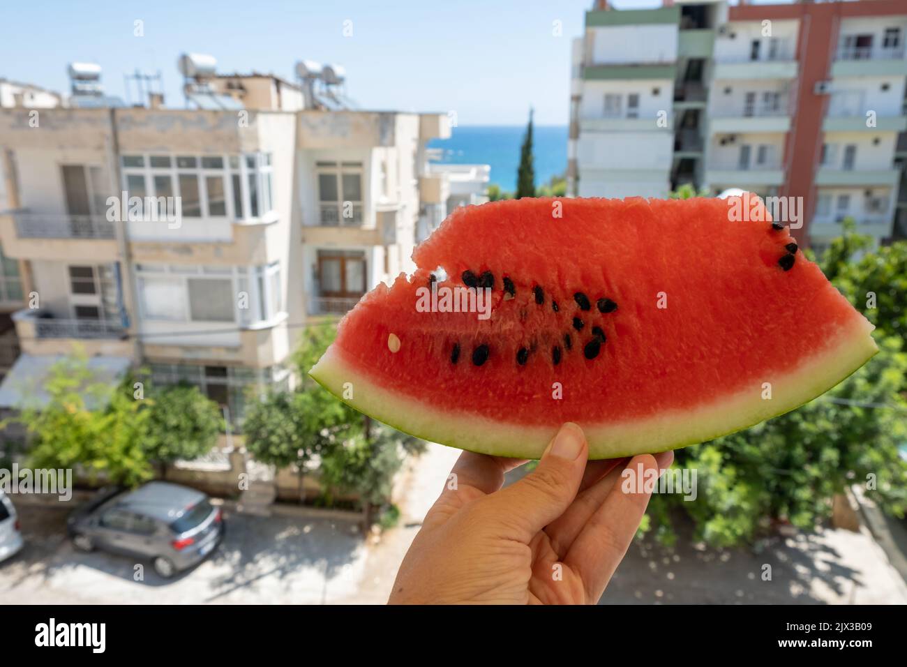 Hand holds piece of watermelon against background of beach town and sea ...