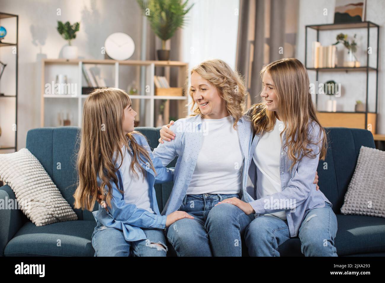Happy blond sisters sitting on couch hug excited young mom or nanny ...