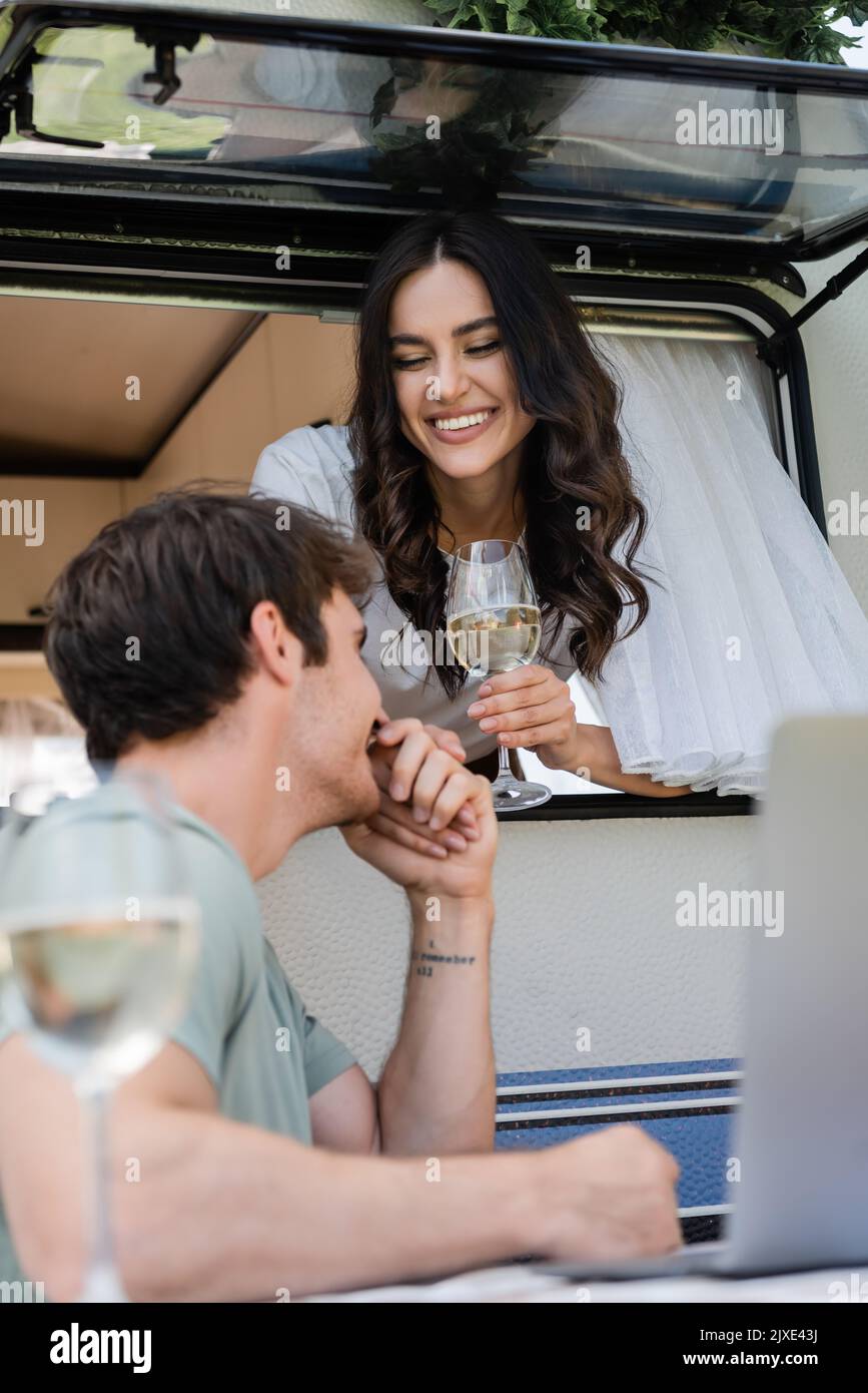 Smiling man kissing hand of girlfriend in camper van near vine and ...