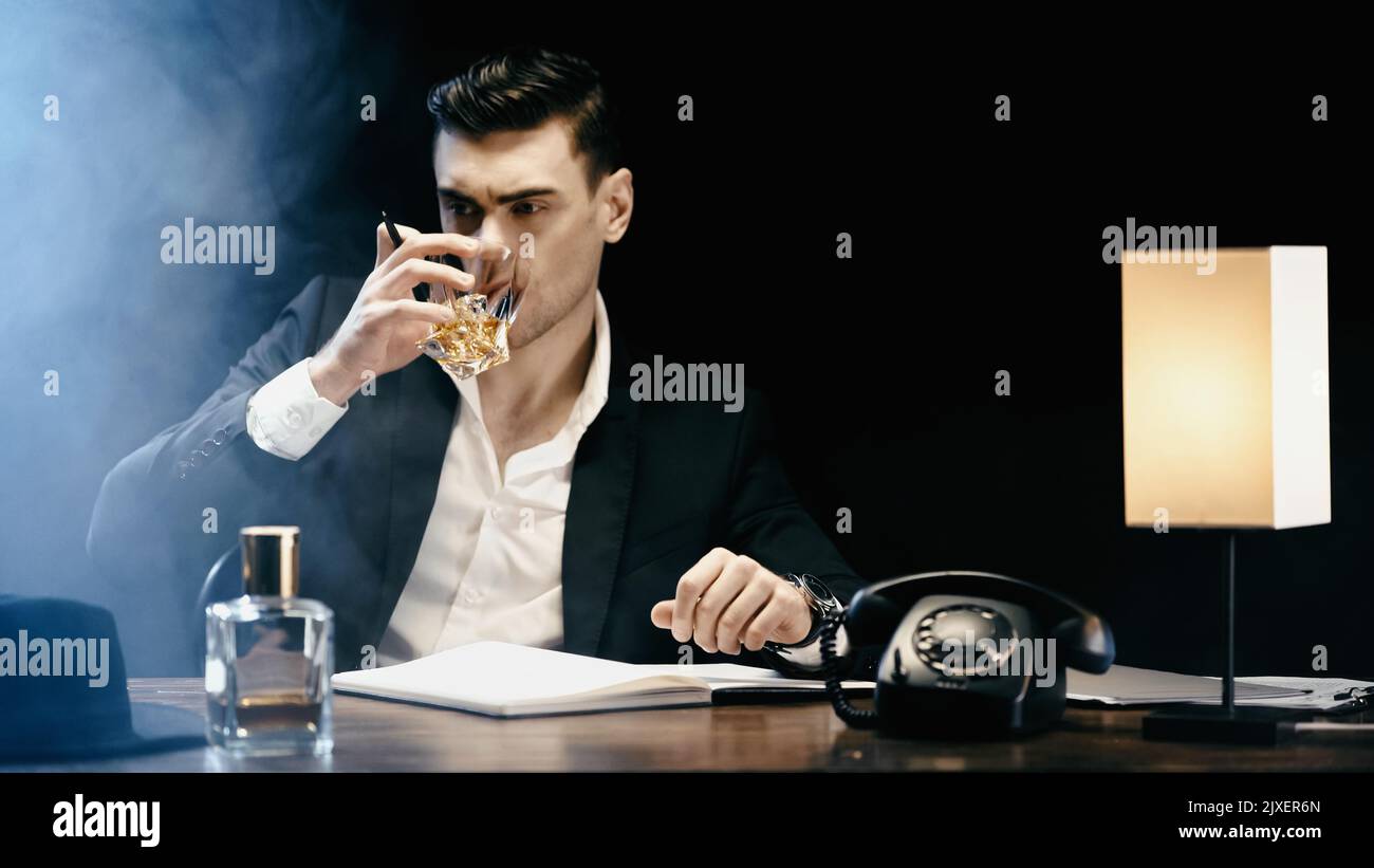 Businessman drinking whiskey near telephone and notebook on black ...