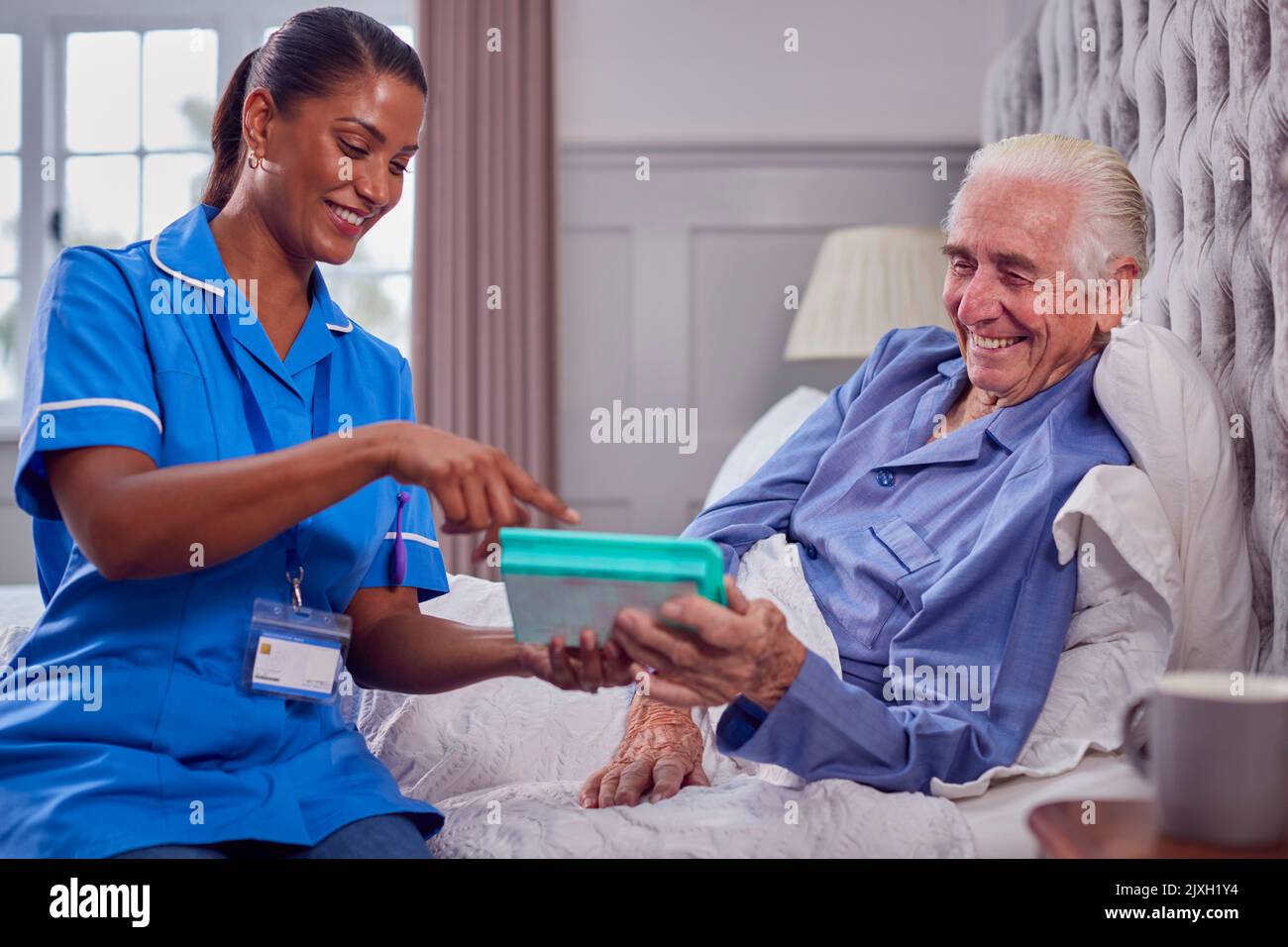 Female Care Worker In Uniform Helping Senior Man At Home In Bed With ...