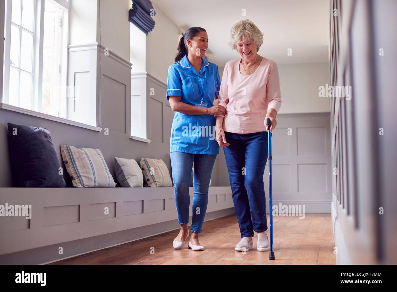 Senior Woman At Home Using Walking Stick Being Helped By Female Care ...