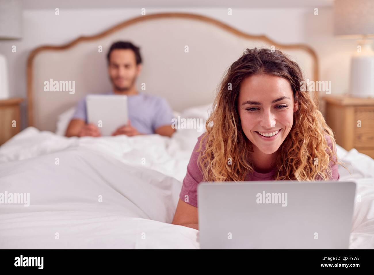 Couple Wearing Pyjamas Lying In Bed At Home With Woman Using Laptop And ...