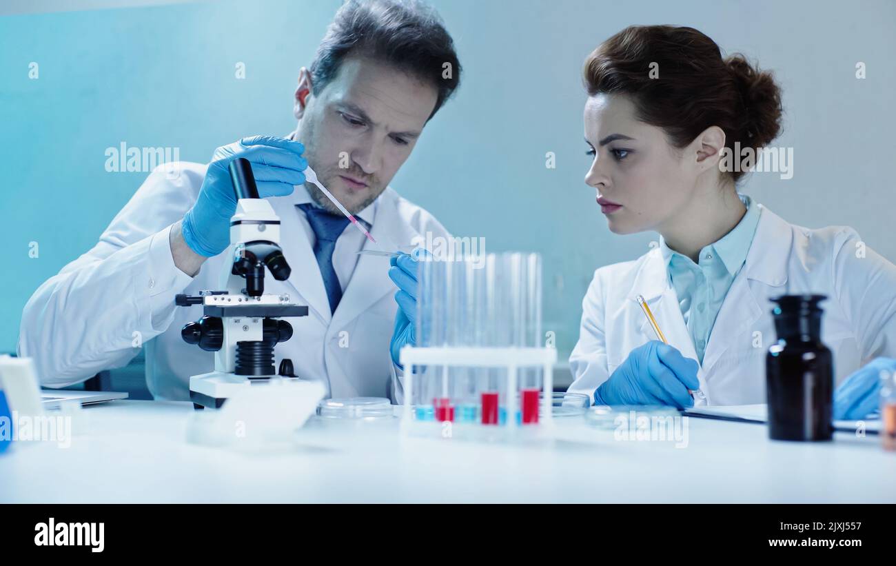 scientist taking sample with pipette near colleague making notes in lab ...