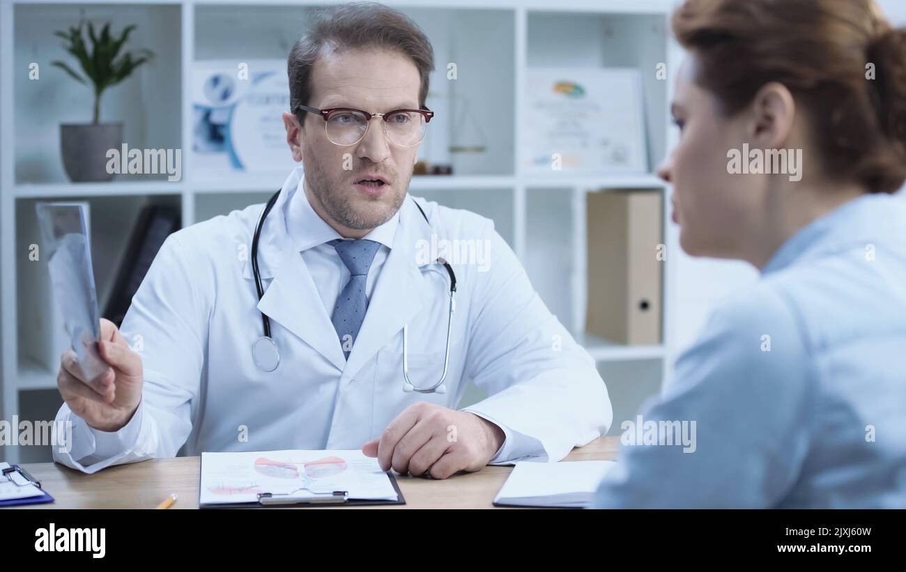 serious doctor holding x-ray scan while talking with patient on blurred ...