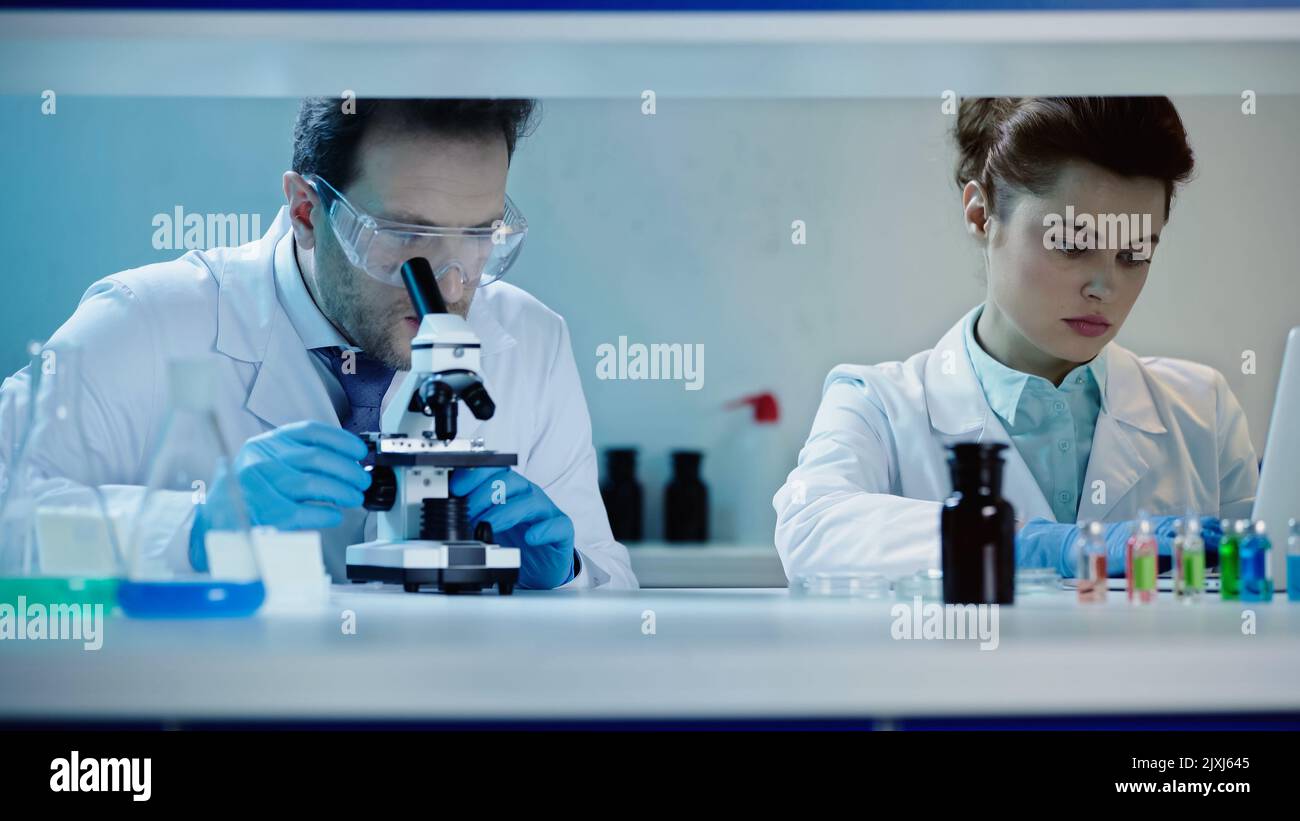 scientist in goggles looking through microscope near colleague using ...